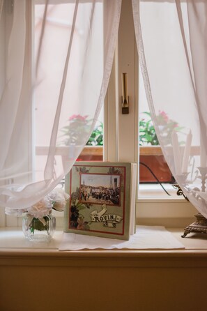Uma janela com cortinas leves, uma fotografia antiga emoldurada e um vaso com flores no parapeito.