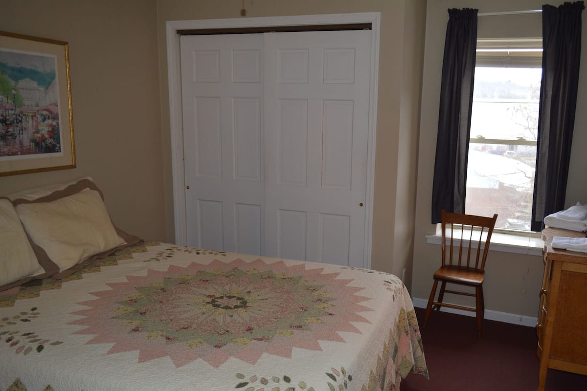 Quarto com cama, cadeira, escrivaninha e janela com cortinas.