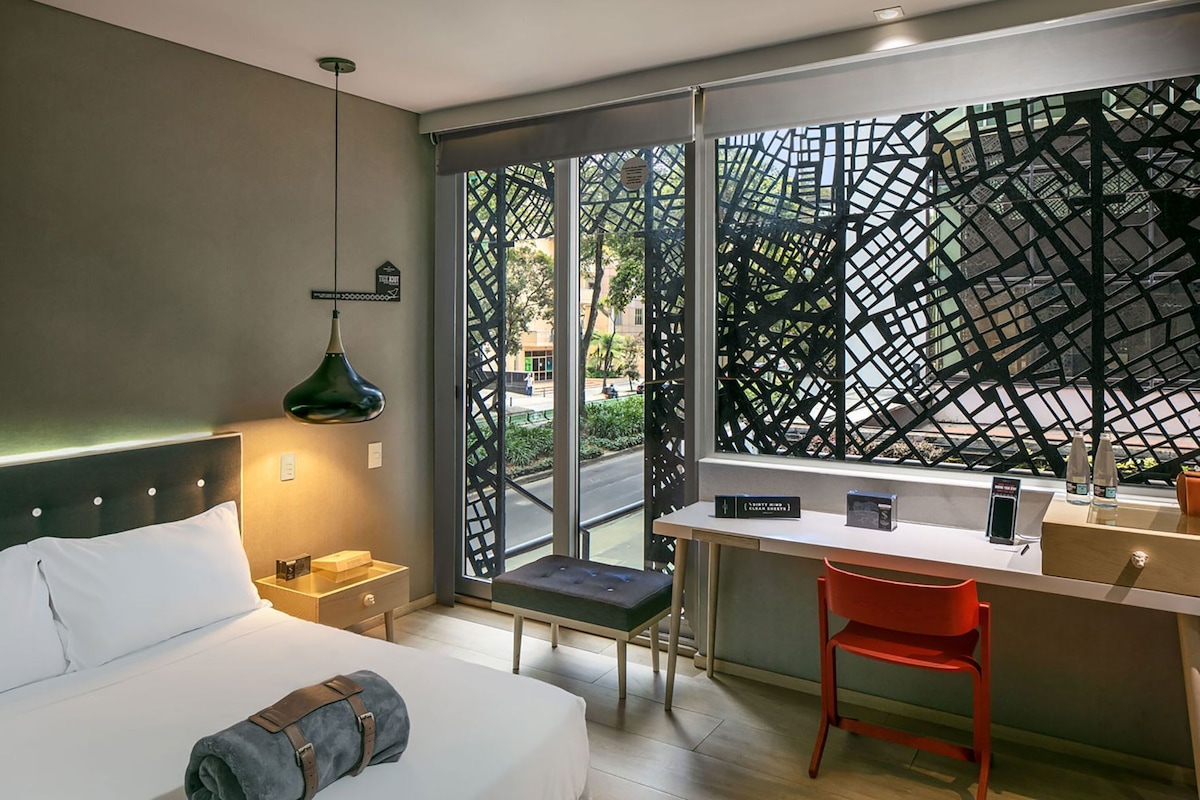 Quarto de hotel com cama, escrivaninha, cadeira e vista pela janela com padrão de treliça.