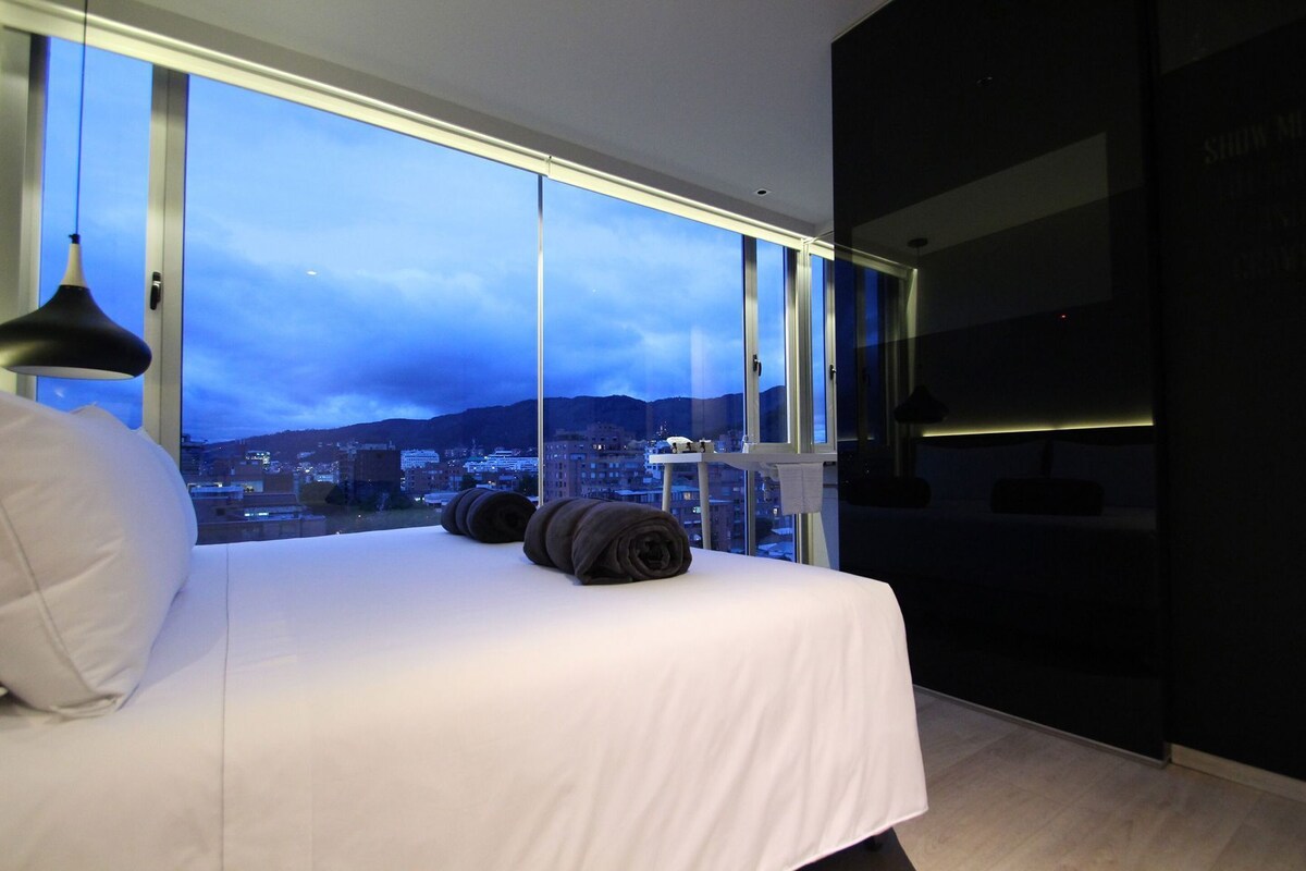 Quarto de hotel moderno com uma cama grande, vista para a paisagem urbana e horizonte visível por grandes janelas.