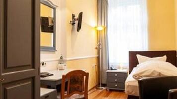 Quarto de hotel com piso de madeira, cama com roupa de cama branca, escrivaninha com cadeira, guarda-roupa e janela com cortinas.
