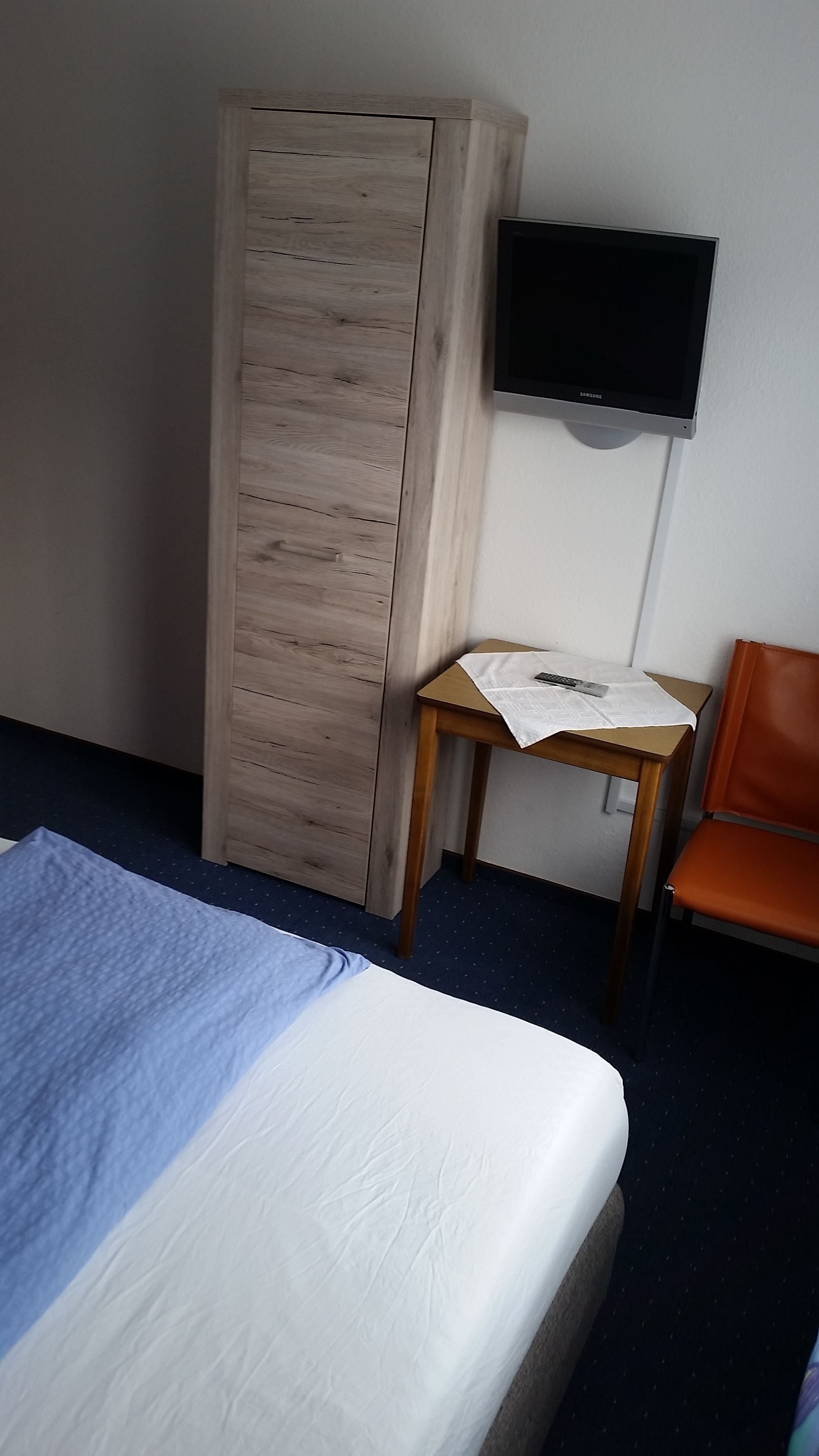 Quarto de hotel com uma cama, guarda-roupa de madeira, uma mesinha com um comando remoto, televisão na parede e uma cadeira laranja.