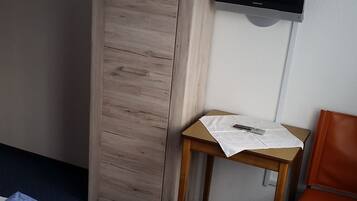 Quarto de hotel com uma cama, guarda-roupa de madeira, uma mesinha com um comando remoto, televisão na parede e uma cadeira laranja.