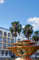 Um hotel com uma fonte em frente, palmeiras e um céu azul límpido.