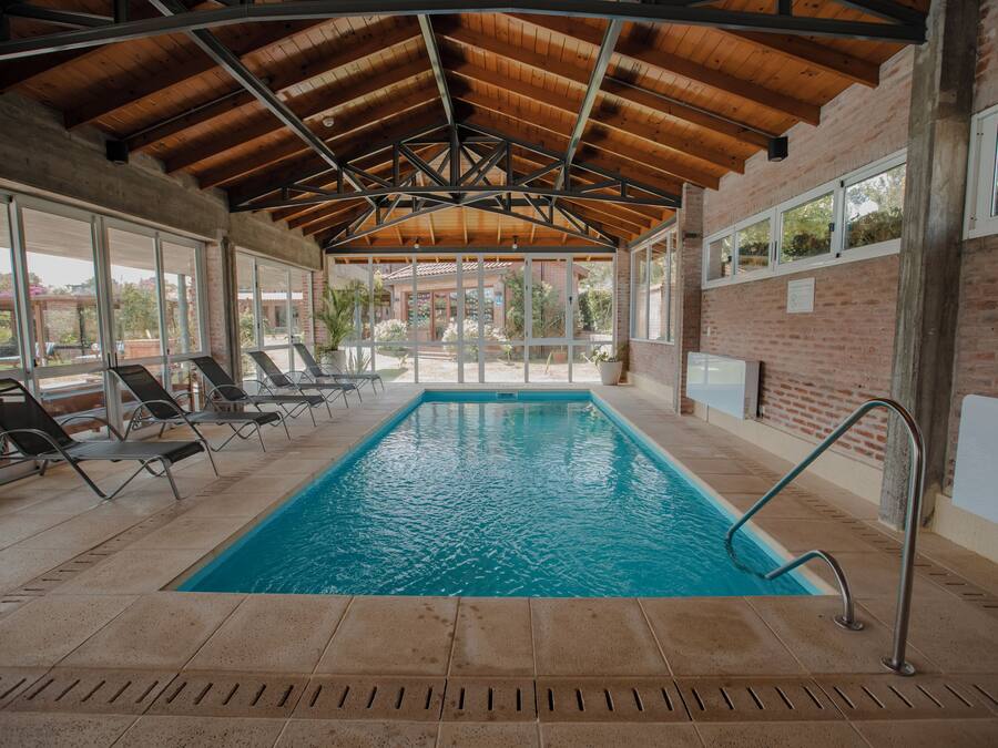 Piscina interna com teto de madeira e chão de azulejos, cercada por espreguiçadeiras e grandes janelas.