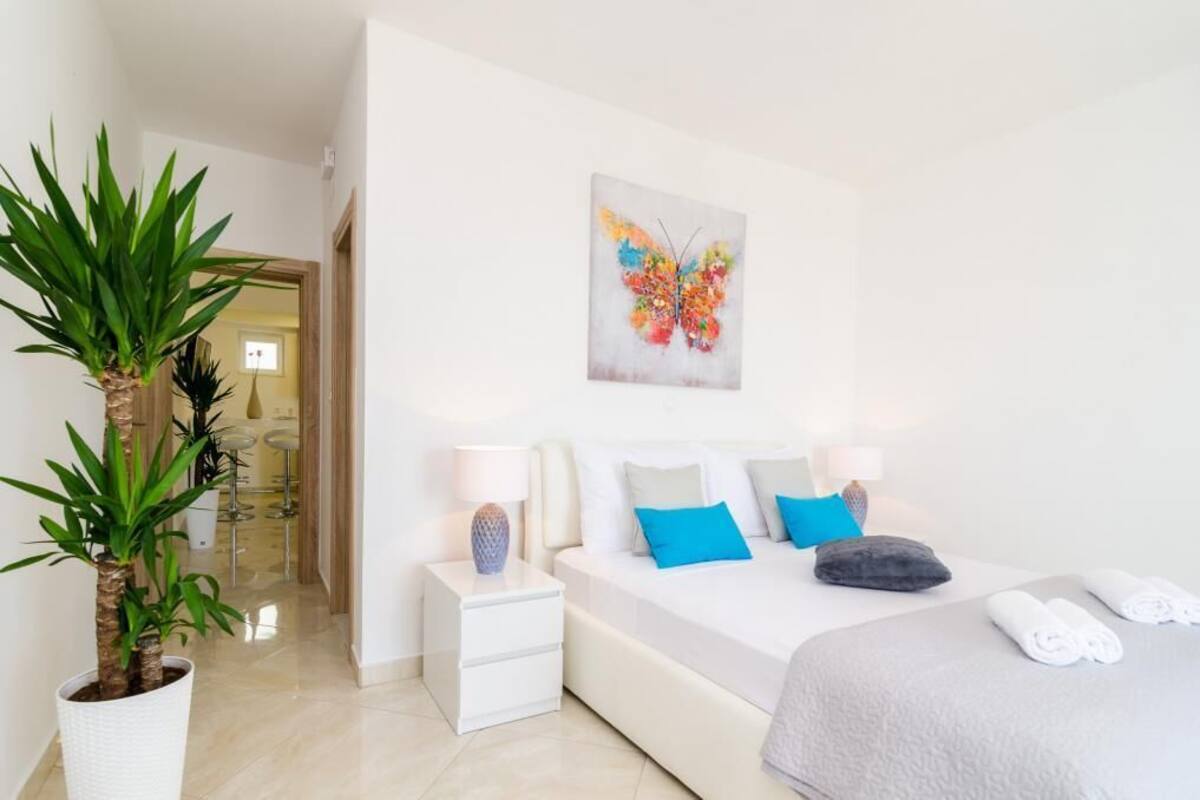 Quarto moderno com uma cama grande, duas mesas de cabeceira, um criado-mudo, uma planta em vaso e um quadro de borboleta colorida na parede.