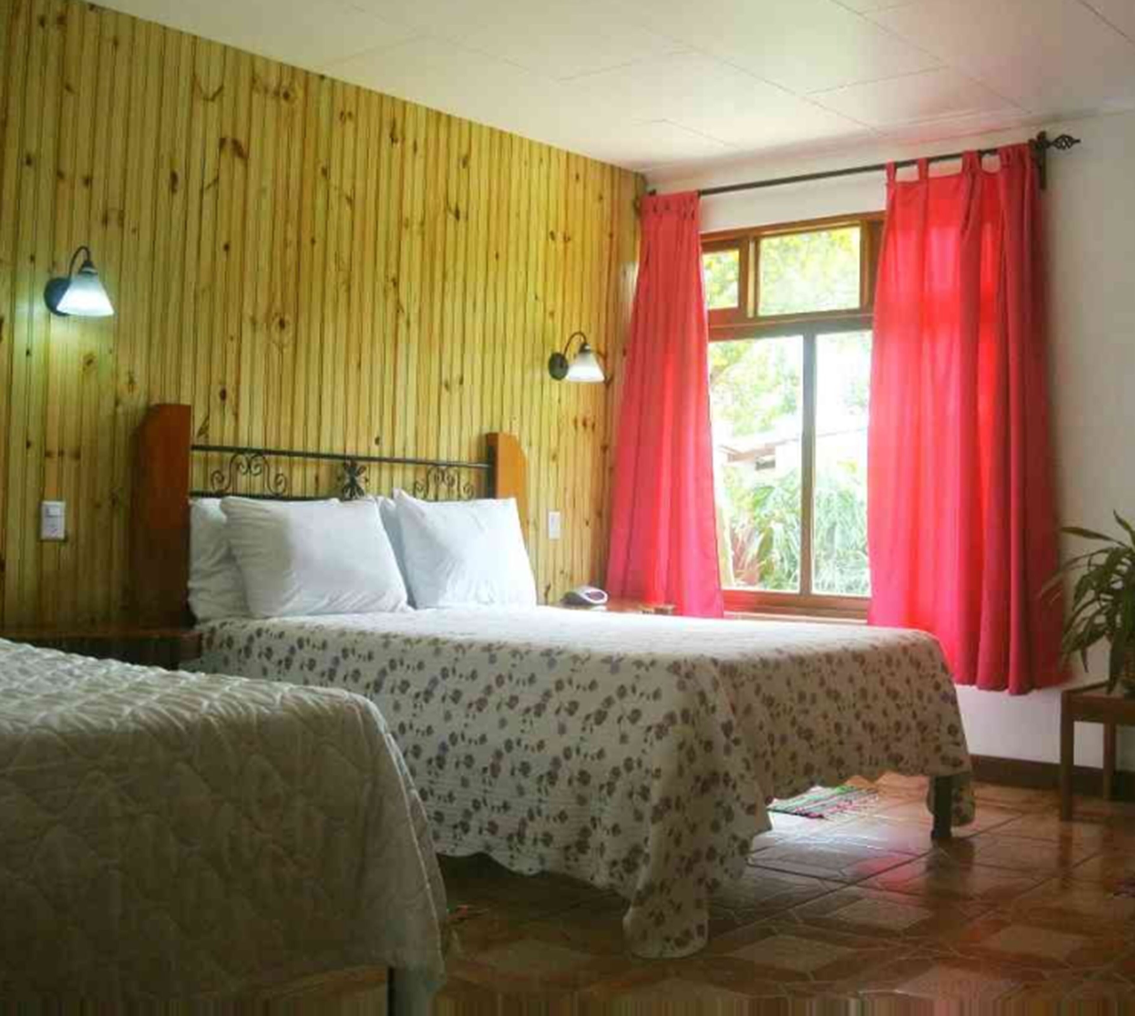 Quarto com duas camas, paredes de madeira, uma janela com cortinas vermelhas e uma pequena mesa.