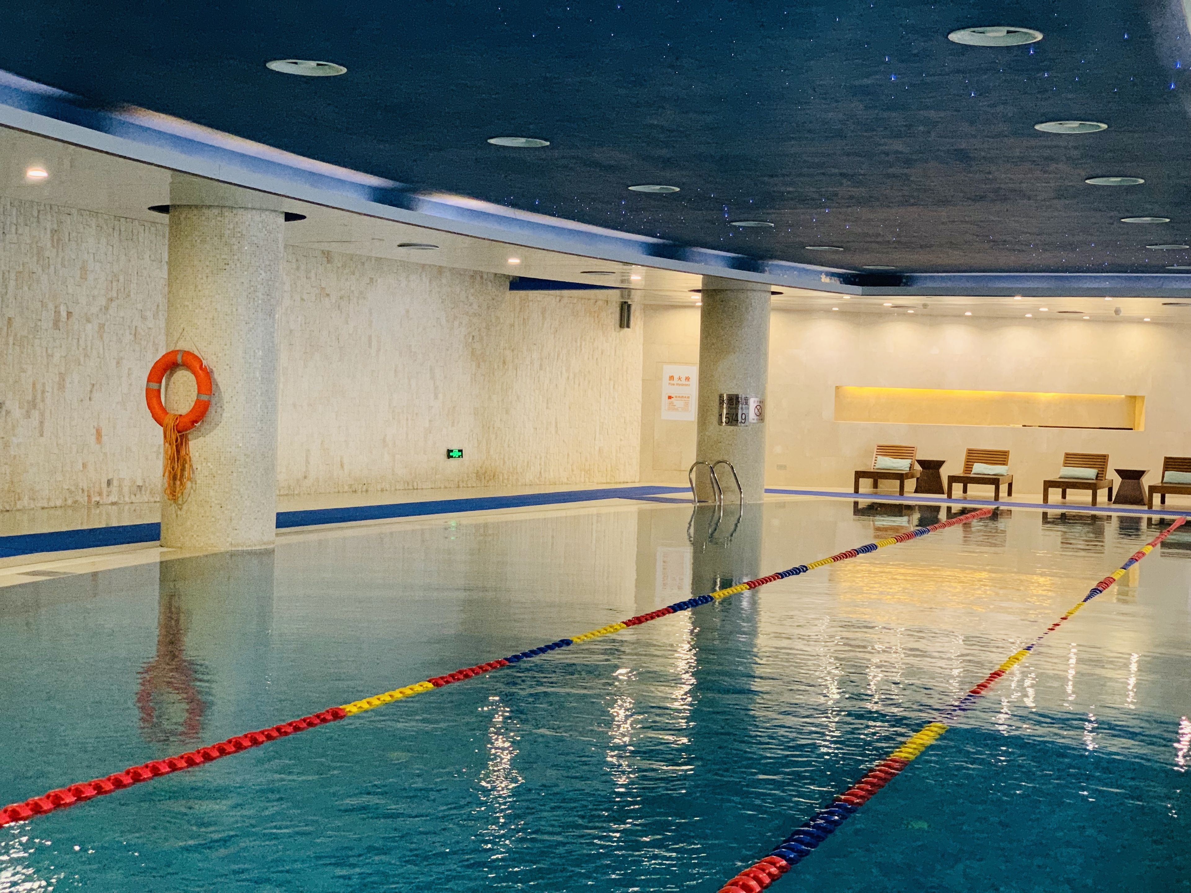 Piscina com divisórias de pista claras, um salva-vidas na parede e uma área de estar com cadeiras.