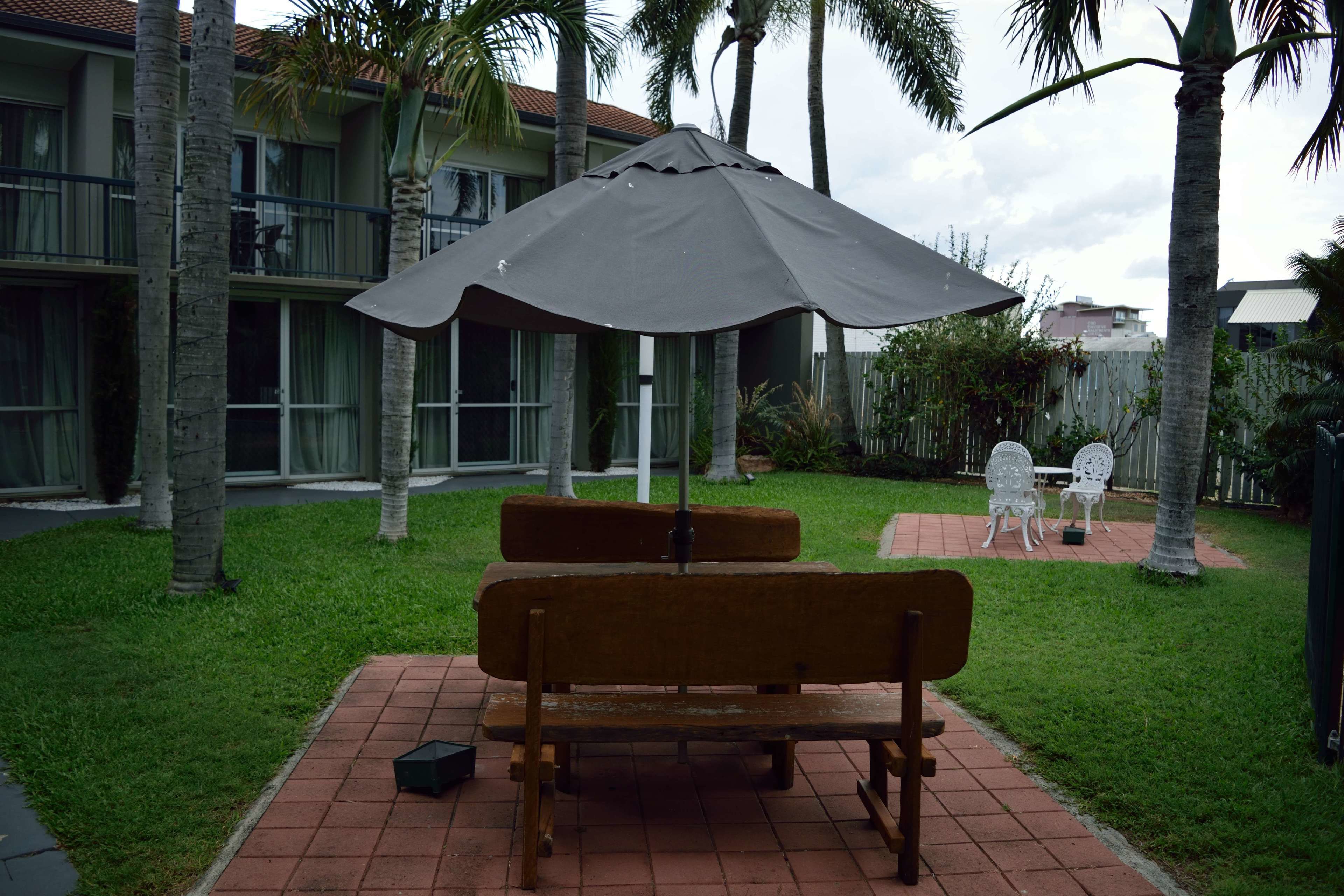 Um banco de madeira sob um grande guarda-sol num pátio com mesa de bilhar e cadeiras, cercado por palmeiras e um edifício com varandas.