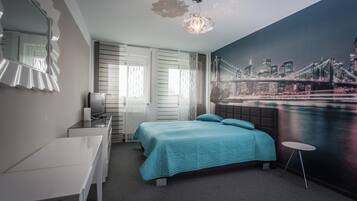 Quarto moderno com cama, escrivaninha e cadeira. Há uma janela grande com persianas e um mural com vista para a cidade na parede.