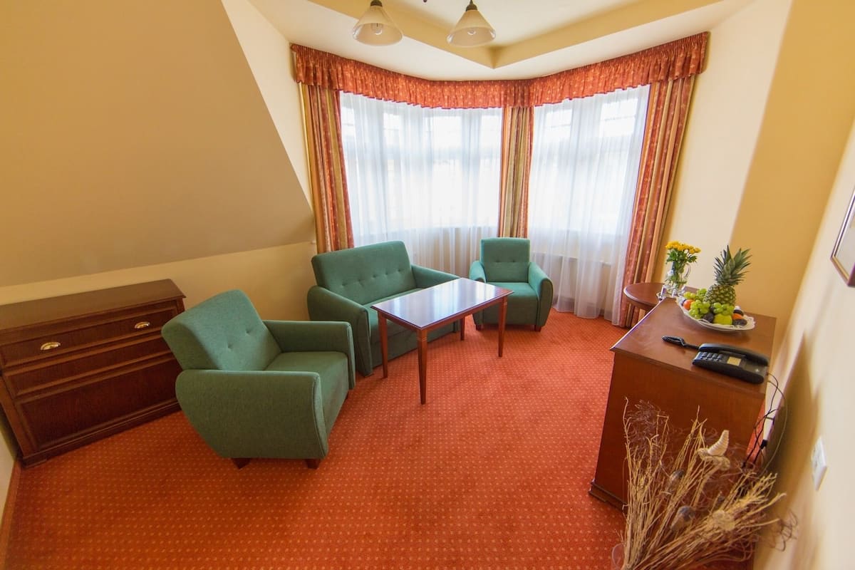 Quarto de hotel com carpete vermelho, duas poltronas verdes, uma mesinha, cômoda de madeira, escrivaninha com telefone e uma fruteira.