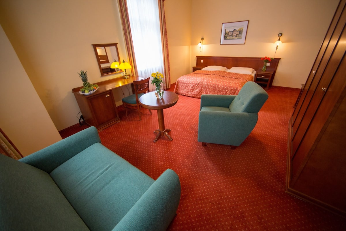 Quarto de hotel com uma cama, duas poltronas, uma mesa pequena com um vaso, uma escrivaninha com abajur e um espelho.