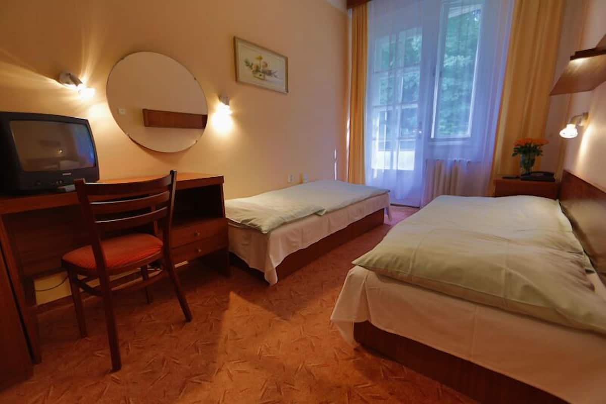 Quarto com escrivaninha de madeira, cadeira, cama, mesinha de cabeceira, abajur, televisão, espelho redondo e janela com cortinas.