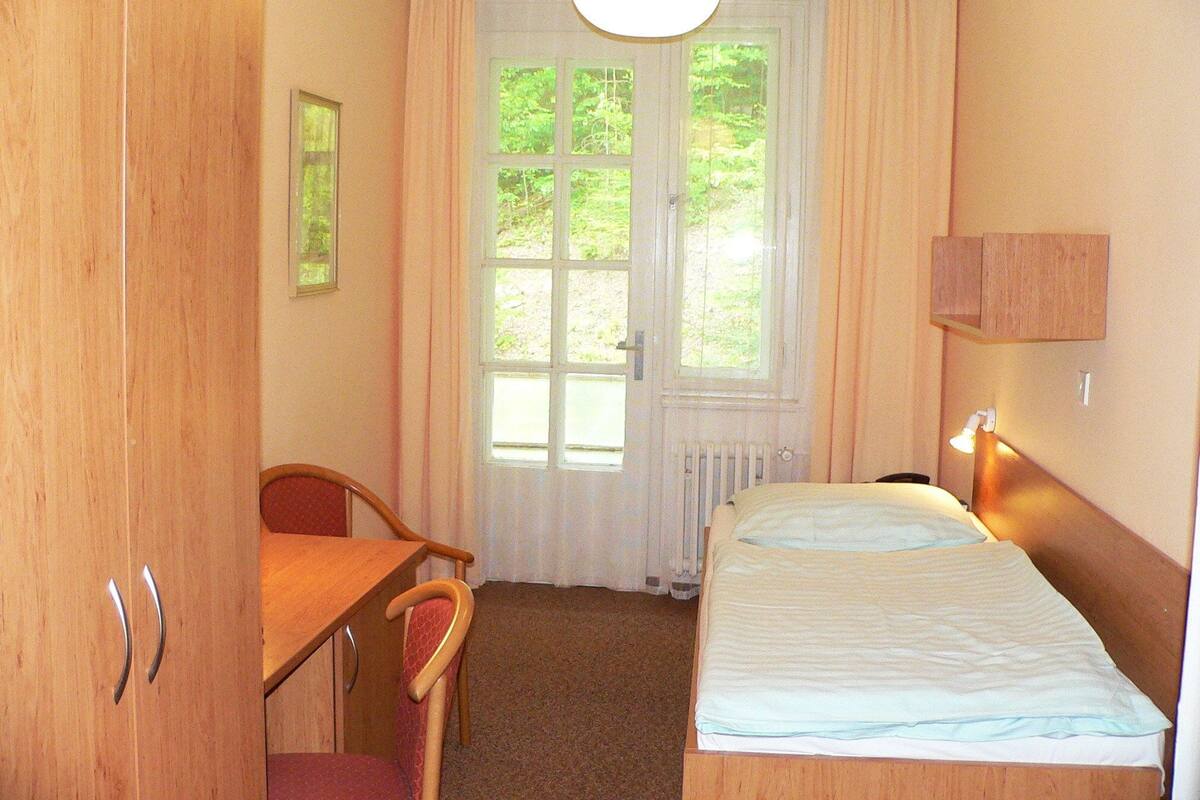 Quarto com cama, escrivaninha e cadeira. Janela com cortinas e vista para o verde lá fora.