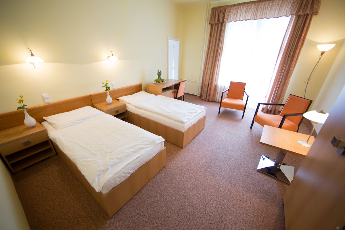 Quarto de hotel com duas camas, uma escrivaninha, uma cadeira, um abajur e uma janela com cortinas.