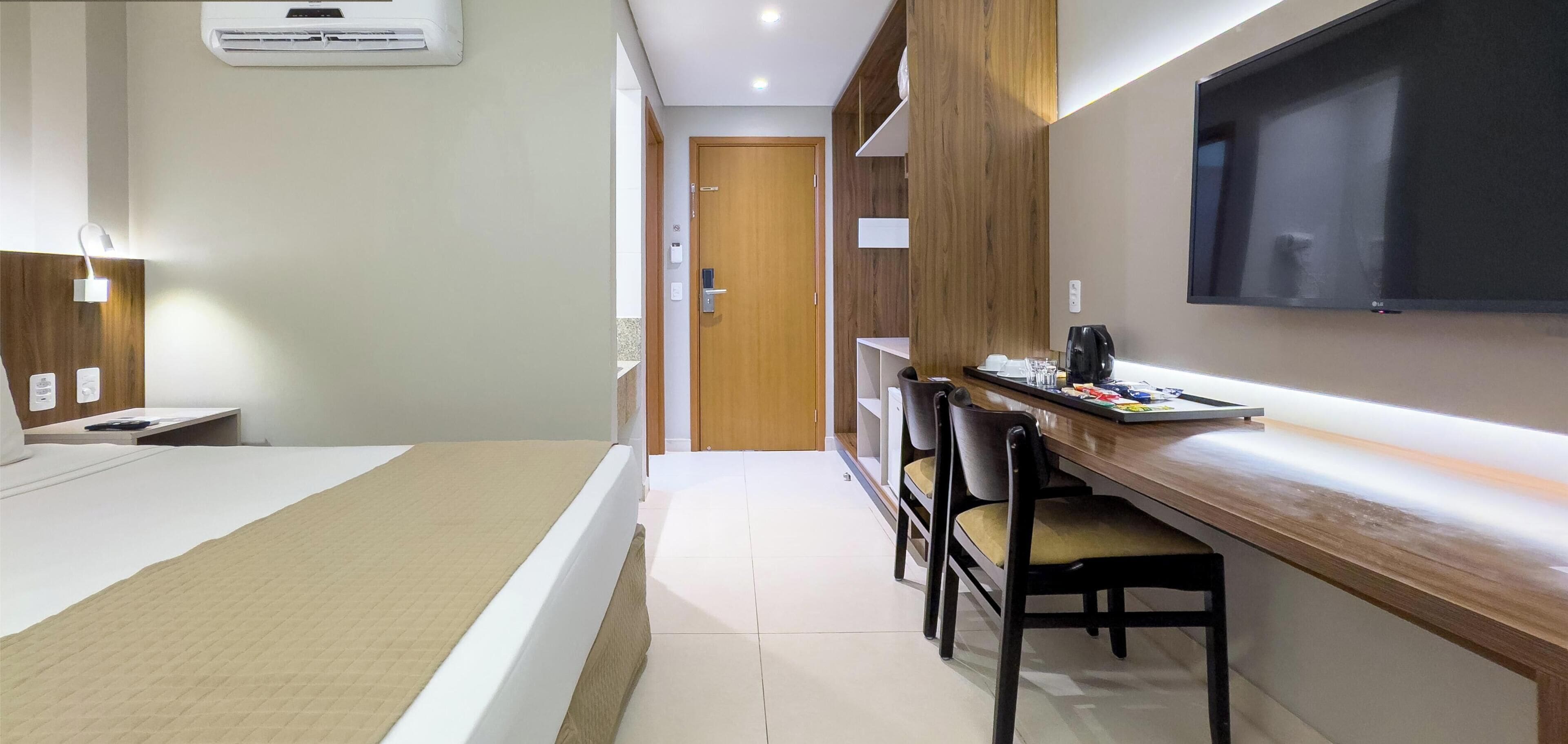 Quarto de hotel com cama, mesa de cabeceira, abajur, televisão, escrivaninha com cadeiras e painel de parede em madeira.