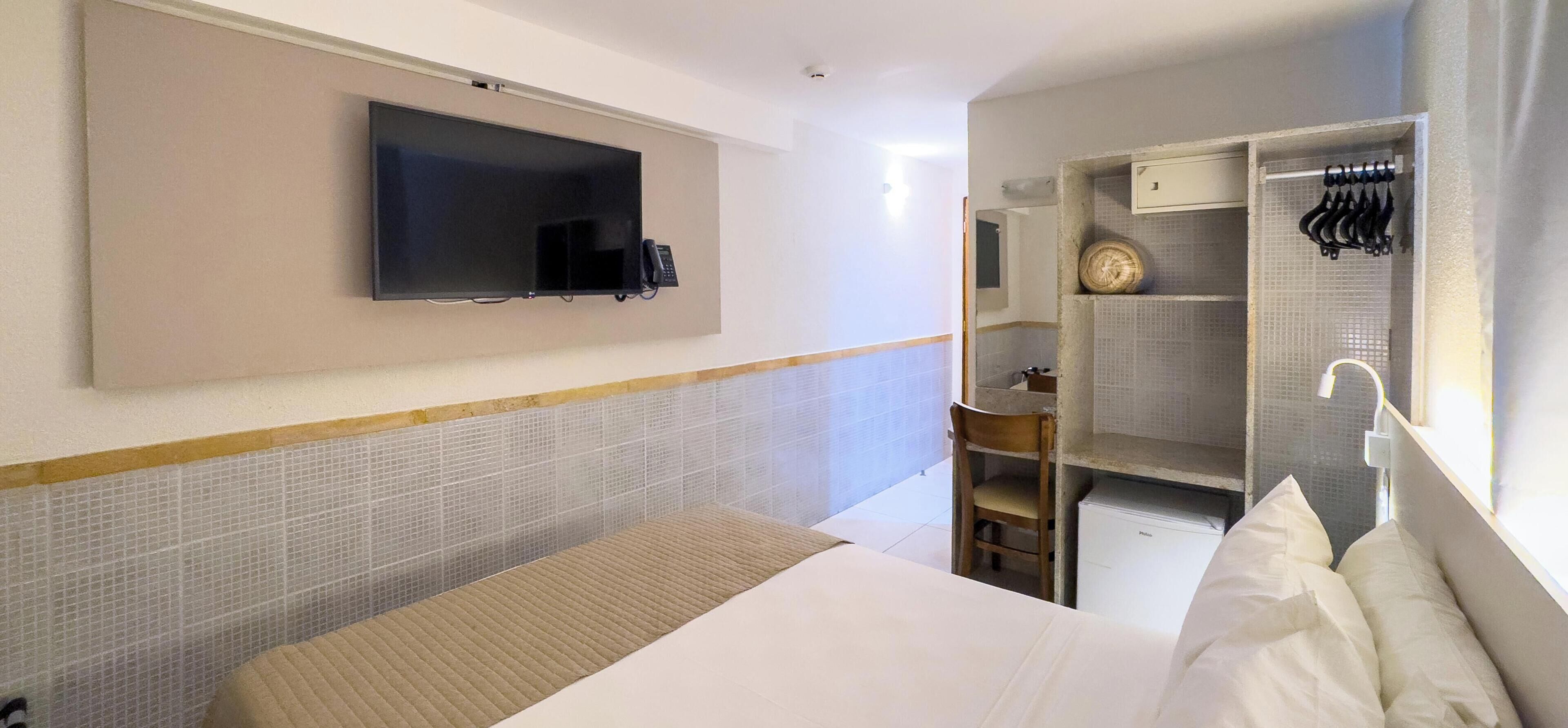 Quarto de hotel com cama, televisão fixada na parede, guarda-roupa com roupas penduradas e uma pequena mesa com cadeira.