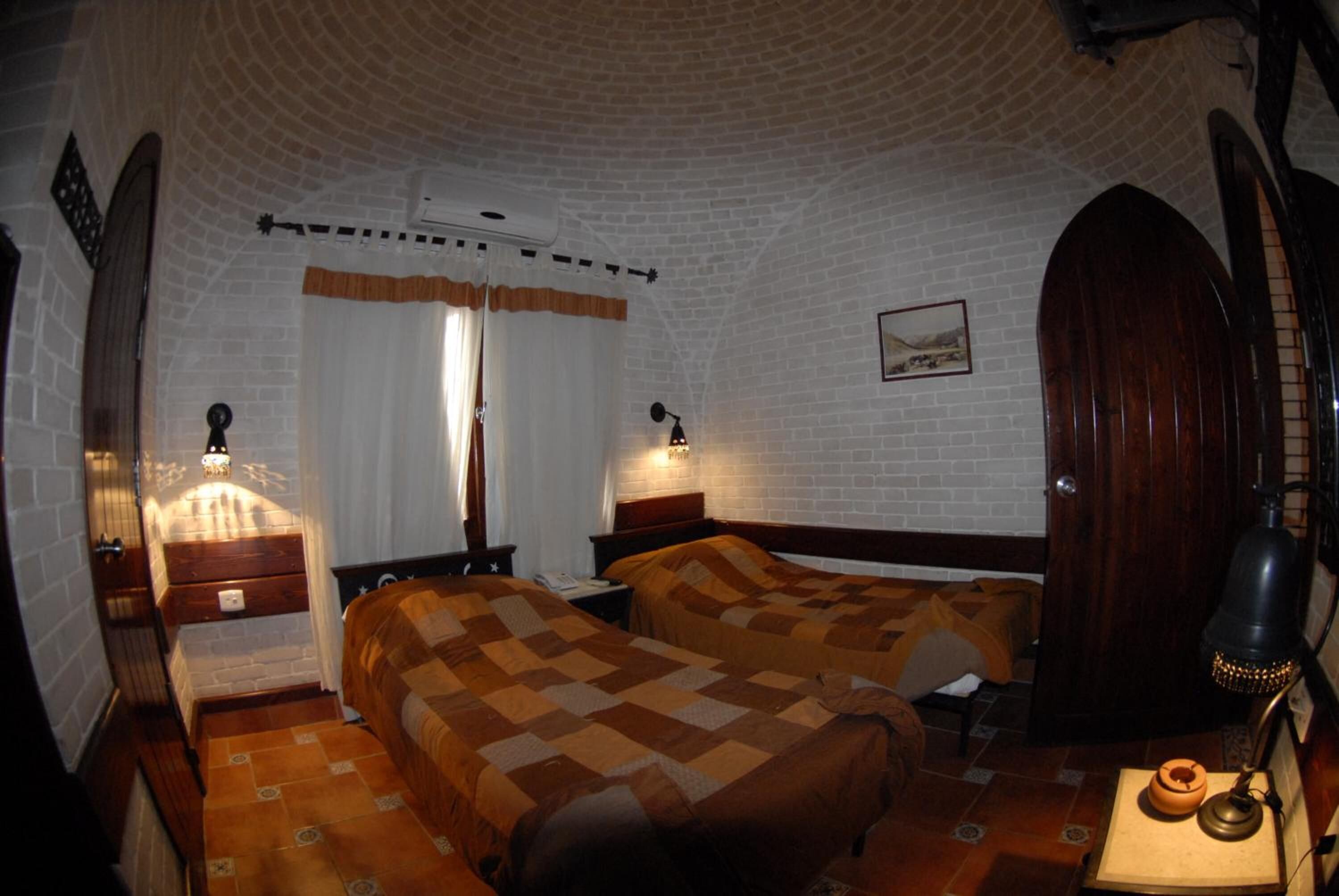 Quarto com duas camas, porta de madeira, parede de tijolos e uma pequena mesa com abajur.