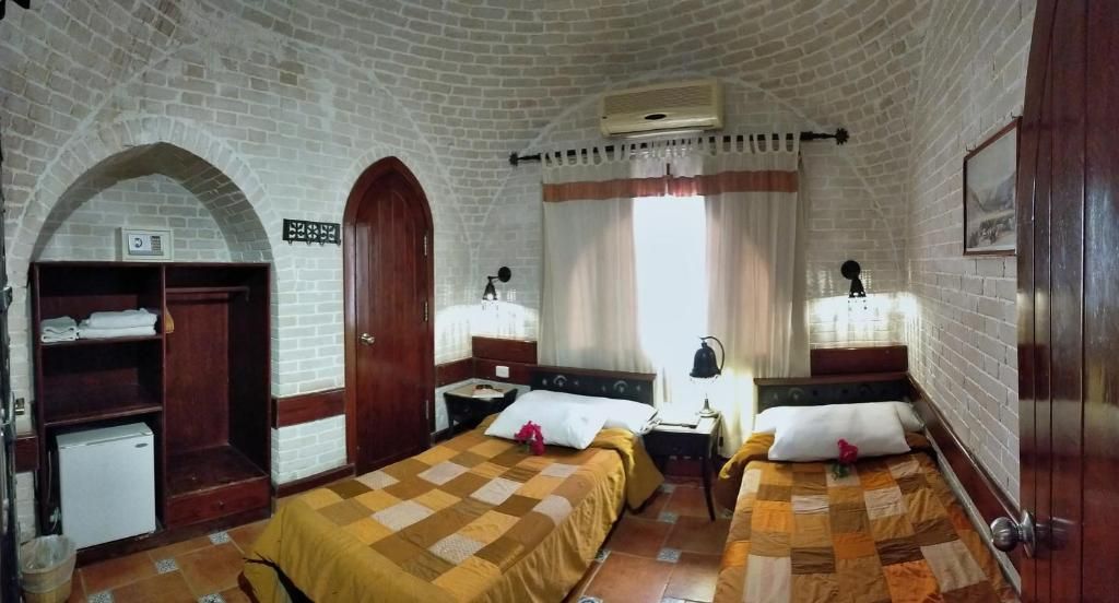 Quarto com duas camas, um guarda-roupa de madeira, um frigorífico pequeno e uma janela com cortinas.