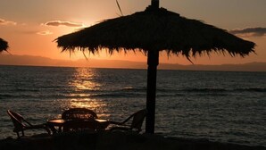 Cena de praia com um guarda-sol de palha, duas cadeiras e uma mesa viradas para o oceano ao pôr do sol.