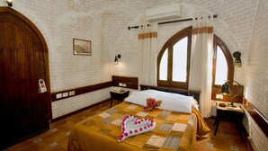 Quarto de hotel com parede de tijolos, porta de madeira, cama grande com decoração em formato de coração, mesa de cabeceira e janela com cortinas.