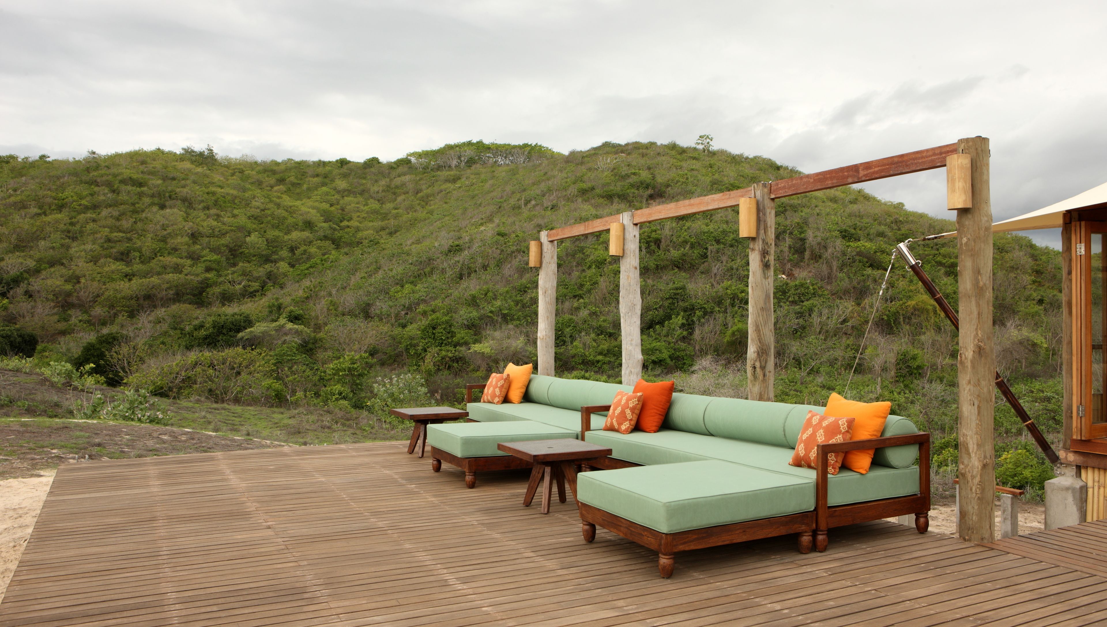 Um deck de madeira com um sofá verde e duas mesas laterais, com um cenário de vegetação exuberante e colinas.