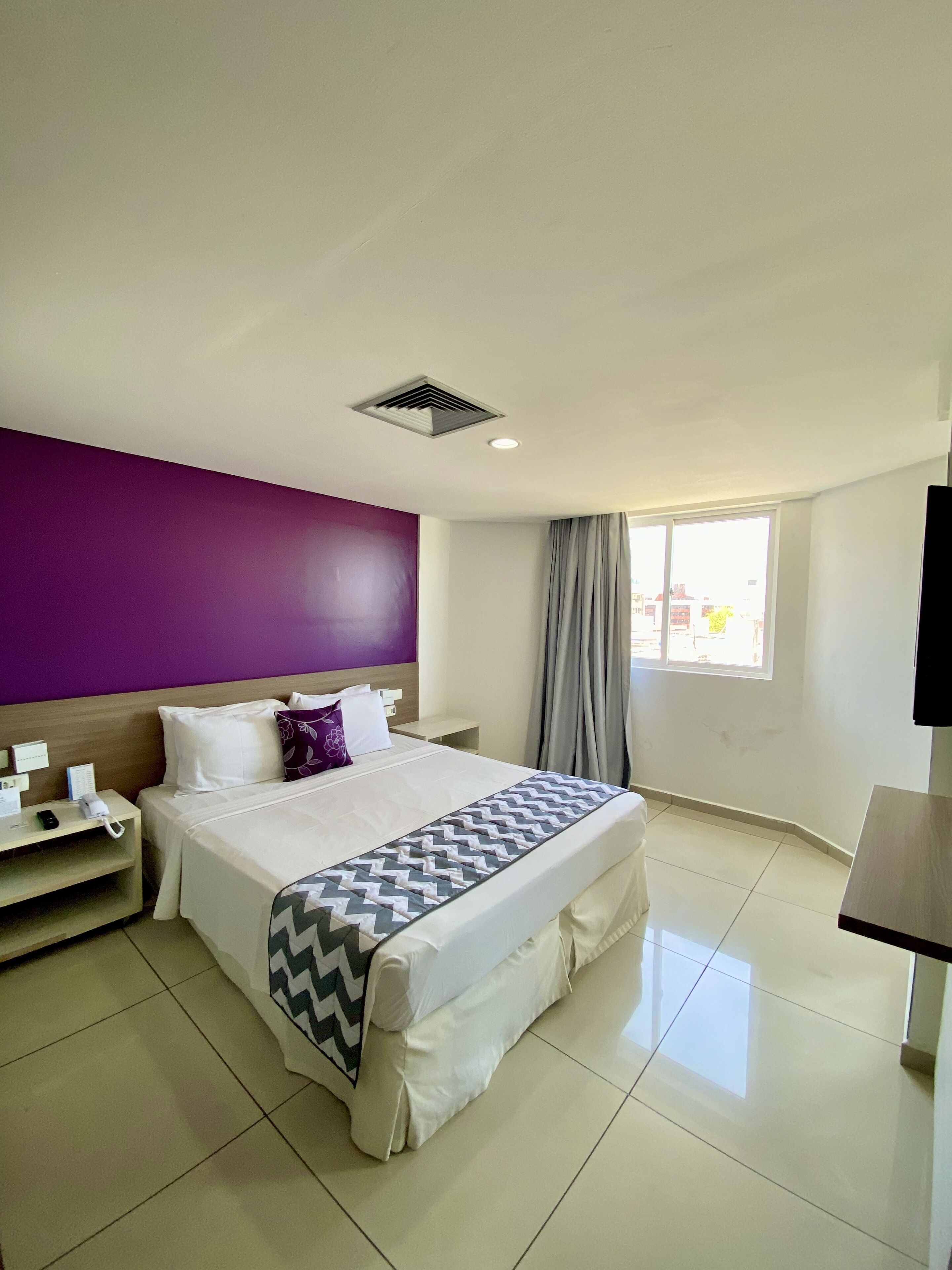 Quarto de hotel com cama, mesa de cabeceira, televisão e janela com cortinas.