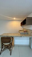 Uma cozinha compacta com bancada de granito, pia embutida e uma cadeira de vime.