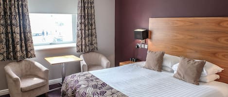 Quarto de hotel com uma cama grande, duas poltronas, uma mesa pequena e uma janela com cortinas.