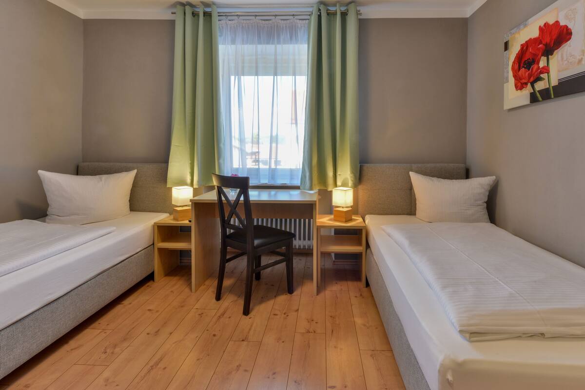 Quarto de hotel com duas camas de solteiro, uma mesinha com uma cadeira e uma janela com cortinas.