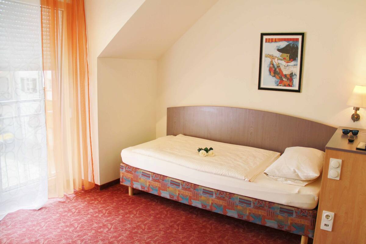 Uma cama de solteiro com colcha estampada, cabeceira, mesa de cabeceira com abajur e um quadro na parede.