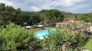 Uma piscina cercada por vegetação exuberante, uma área de pátio com mobiliário de exterior e vista para um morro com árvores e um edifício.