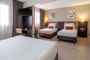 Quarto de hotel com uma cama grande, duas camas menores, uma escrivaninha com cadeira, televisão de tela plana e obras de arte nas paredes.