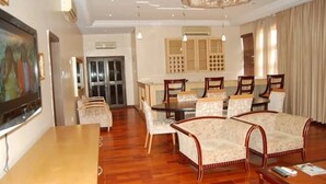 Ampla sala de estar com mesa de jantar de madeira, cadeiras estofadas, televisão e uma grande janela com cortinas.