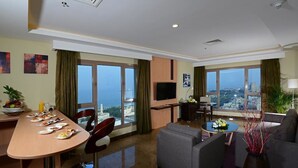 Quarto de hotel com área de jantar, sofá, televisão e grandes janelas com vista para a cidade.