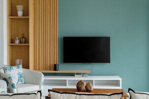 Uma sala de estar moderna com uma TV de tela plana fixada em uma parede verde-água, prateleiras de madeira, um sofá branco com almofadas estampadas e uma mesa de centro de madeira.