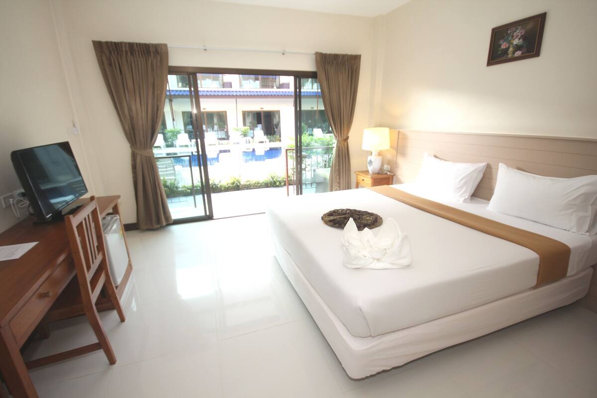 Quarto de hotel com cama, escrivaninha com computador, luminária e vista para uma piscina e um edifício pela janela.