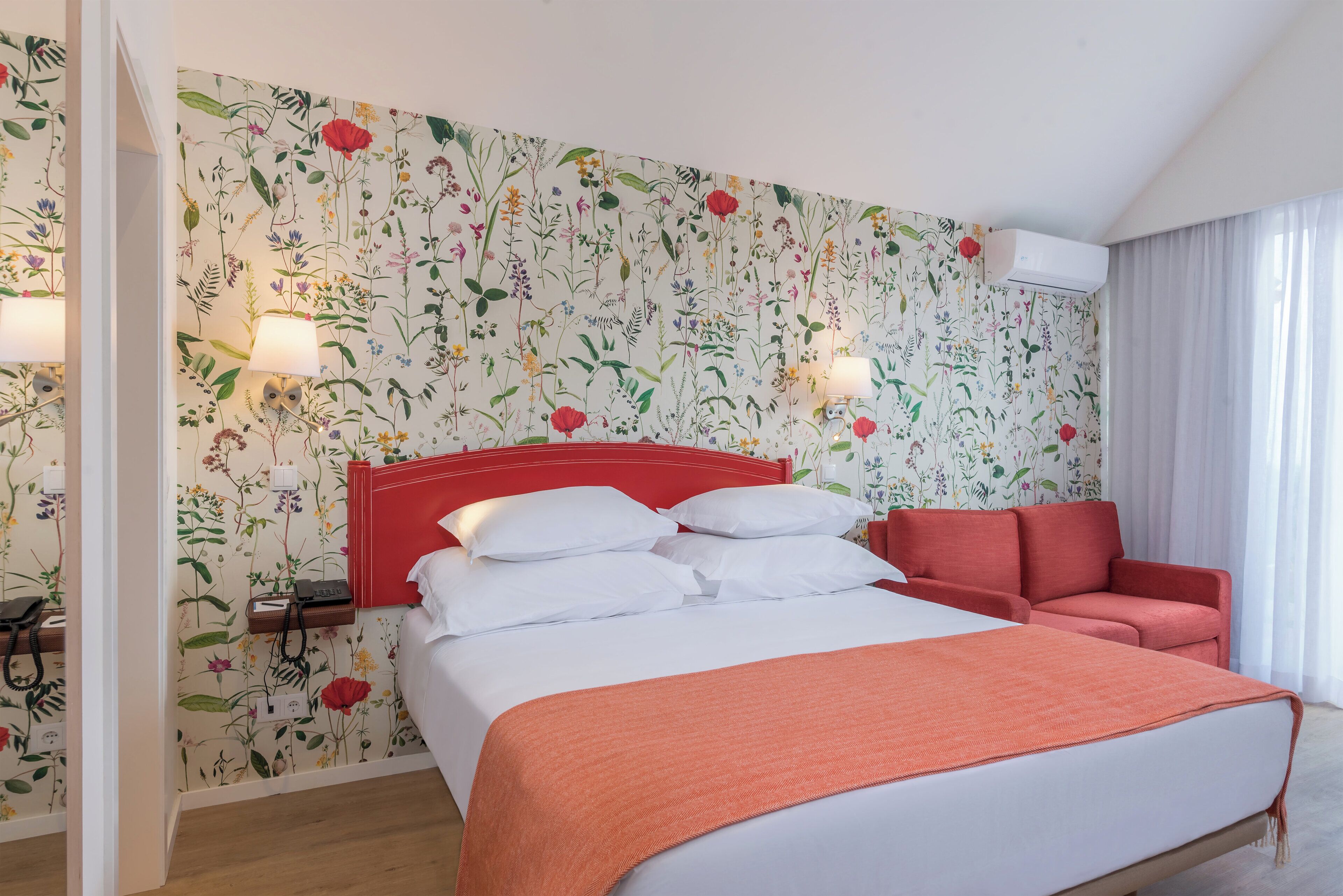 Quarto de hotel com papel de parede floral, cabeceira vermelha, cama com lençóis brancos, sofá vermelho e abajur.