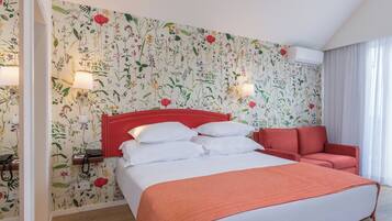 Quarto de hotel com papel de parede floral, cabeceira vermelha, cama com lençóis brancos, sofá vermelho e abajur.