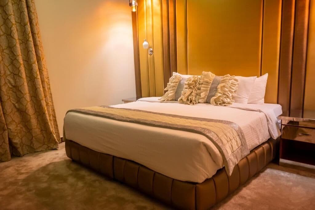 Quarto de hotel com uma cama grande, mesa de cabeceira, abajur e cortina.