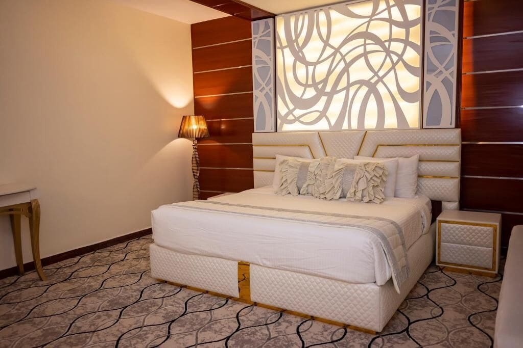 Quarto de hotel com uma cama grande, uma mesa de cabeceira, uma luminária e um painel decorativo na parede.