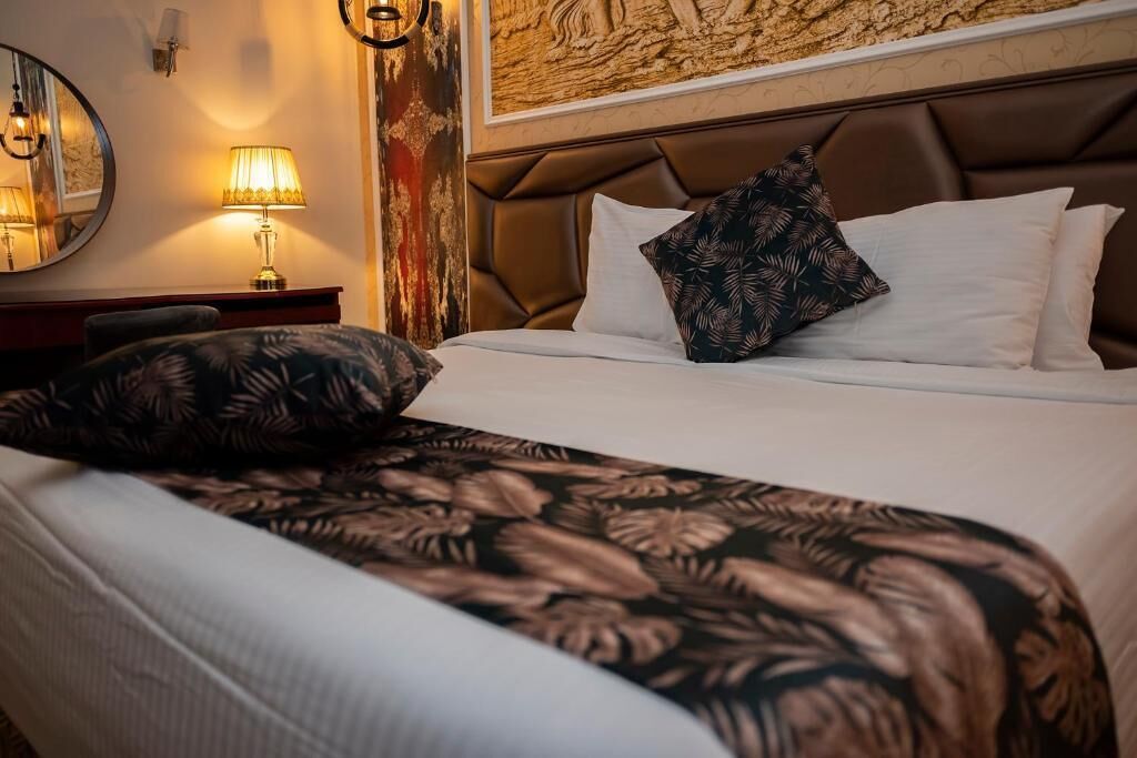 Quarto de hotel com cama, mesa de cabeceira, abajur, espelho e parede texturizada.