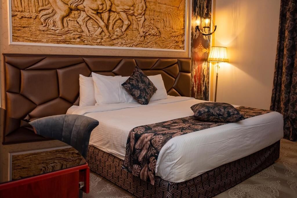 Quarto de hotel com uma cama grande, cabeceira, luminária fixada na parede e mesa de cabeceira vermelha.