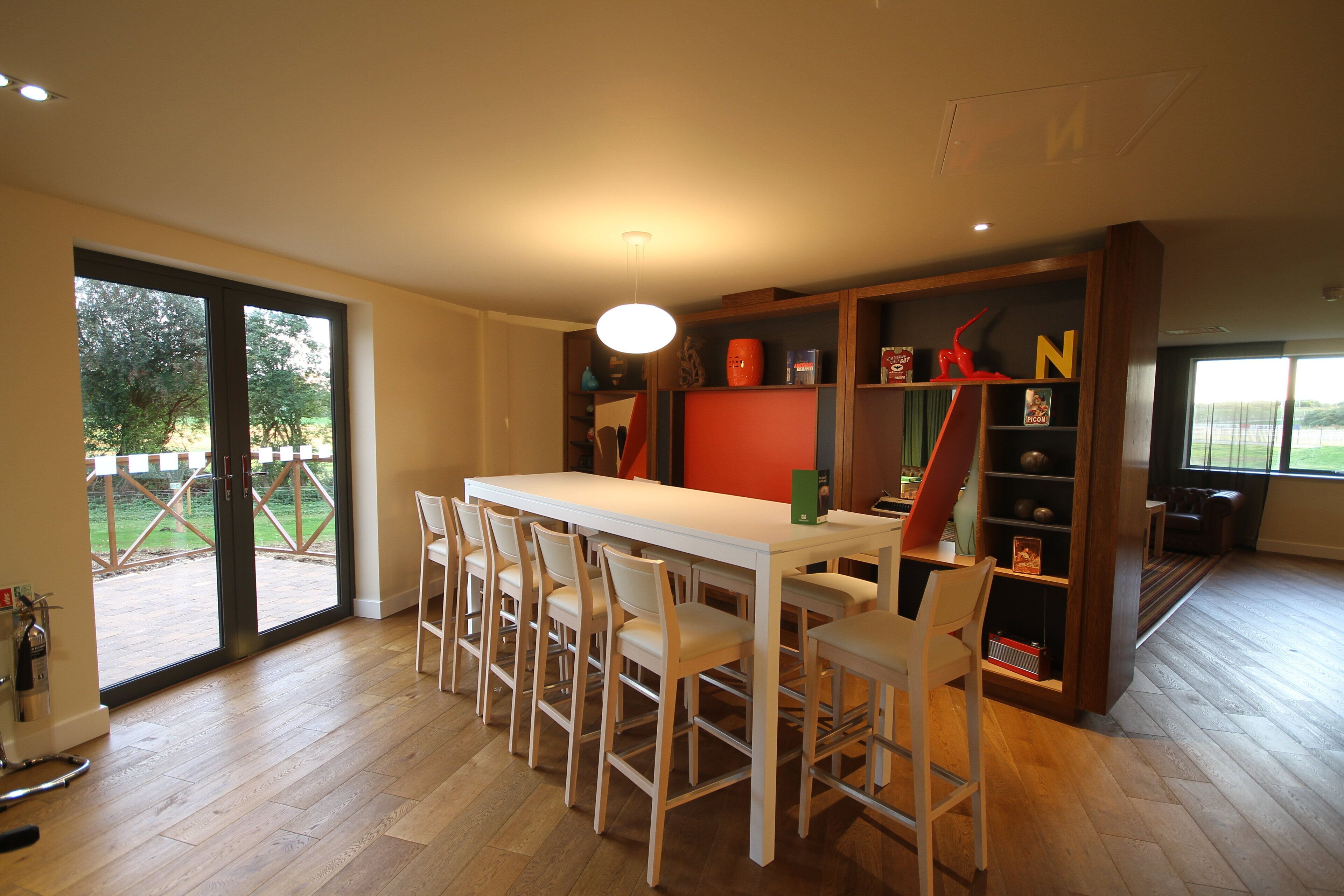 Uma sala de jantar com uma mesa e cadeiras brancas, uma estante com diversos objetos, e vista através de uma porta de vidro.