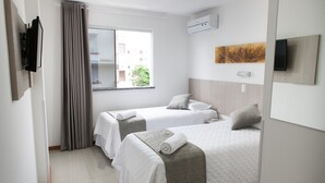 Quarto de hotel com duas camas, uma janela com cortinas, um aparelho de ar condicionado, uma TV de tela plana e uma obra de arte pendurada na parede.