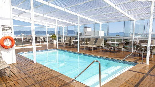 Uma área de piscina ensolarada com vista clara para o mar e montanhas ao fundo.