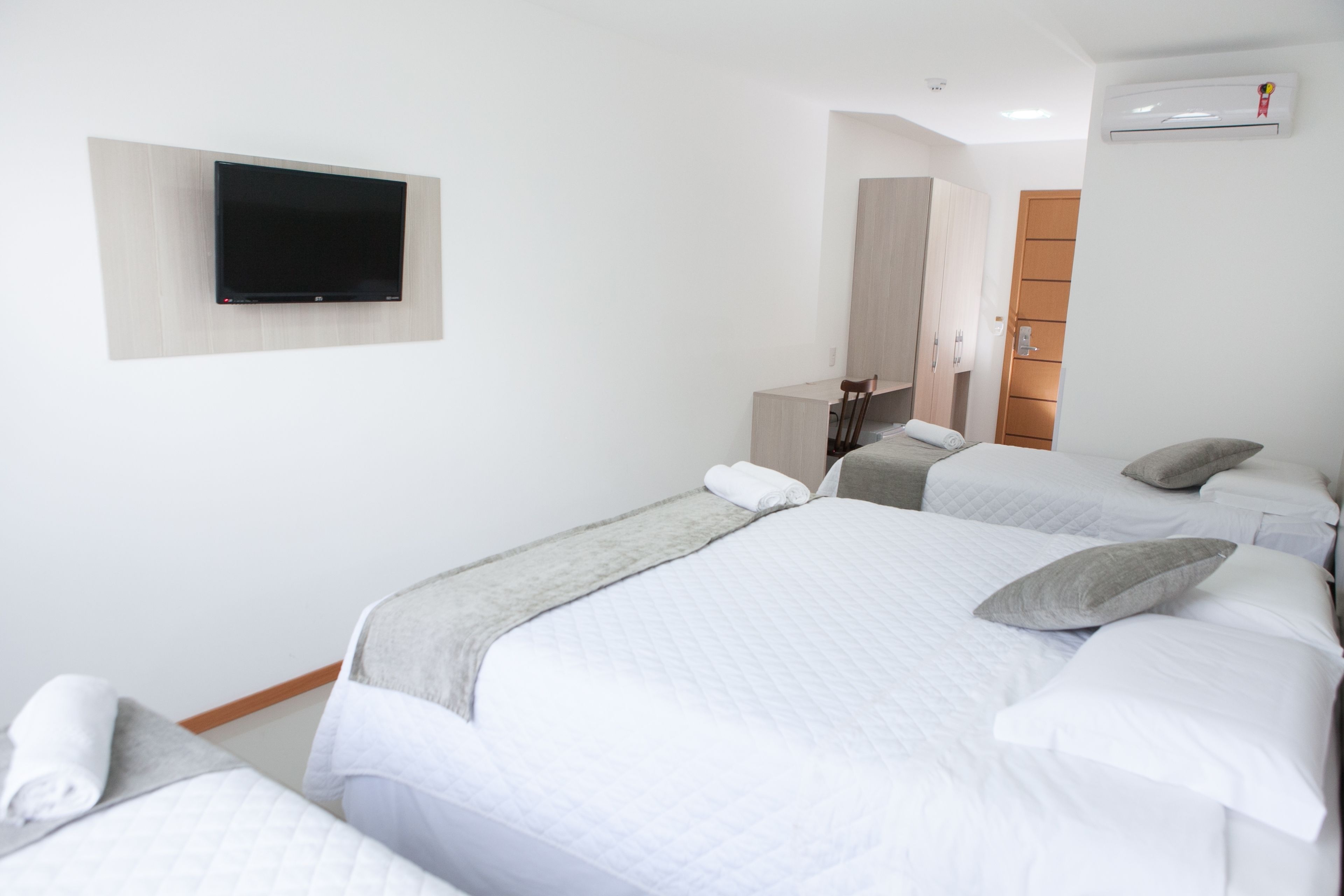 Quarto de hotel com duas camas, uma televisão de tela plana fixada na parede, um guarda-roupa de madeira e um ar condicionado embutido.