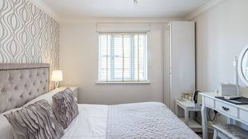 Quarto com cama, mesa de cabeceira com abajur, janela com persianas, escrivaninha com cadeira e espelho.