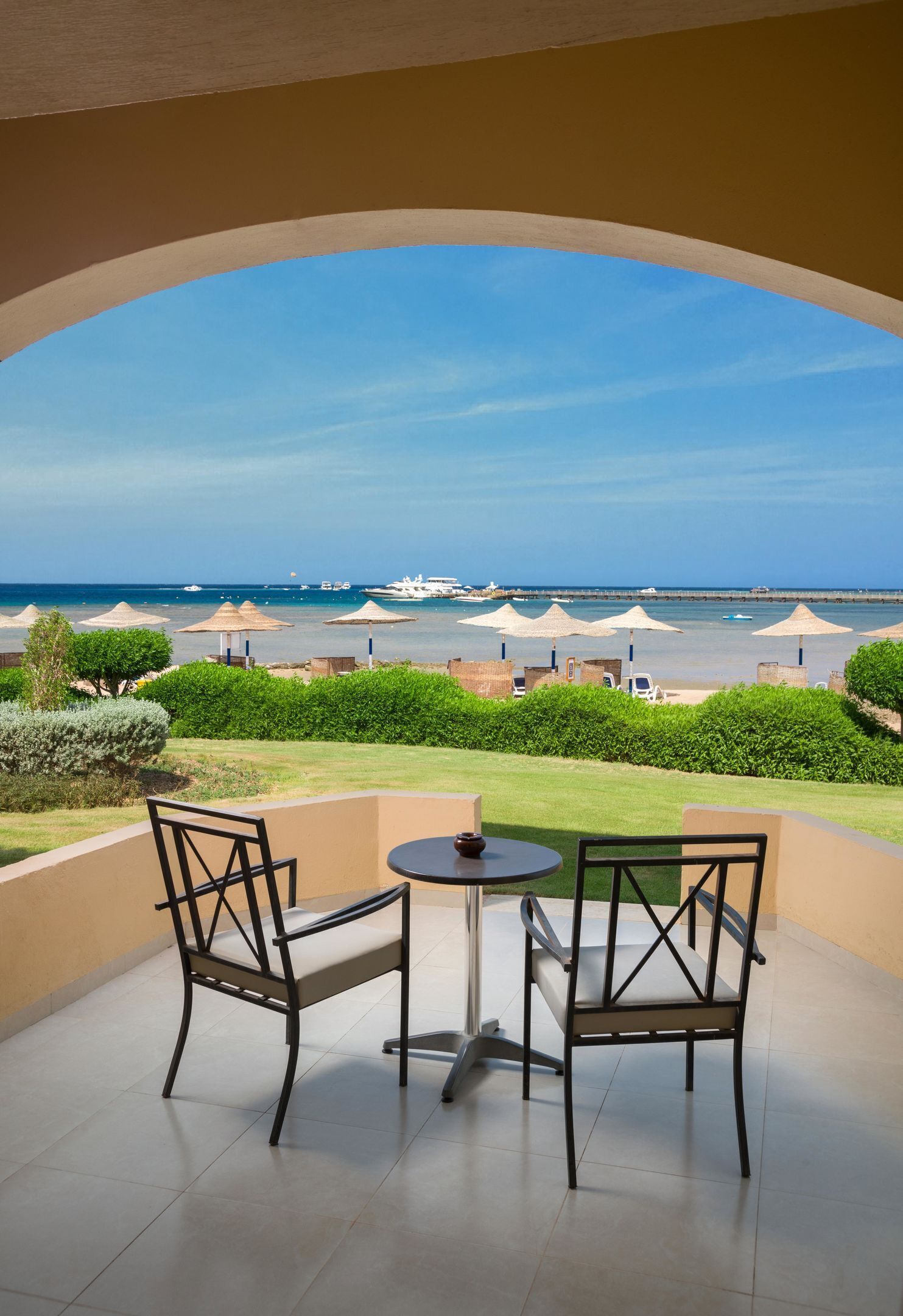Varanda com duas cadeiras e uma mesa, com vista para uma praia com guarda-sóis e barcos.
