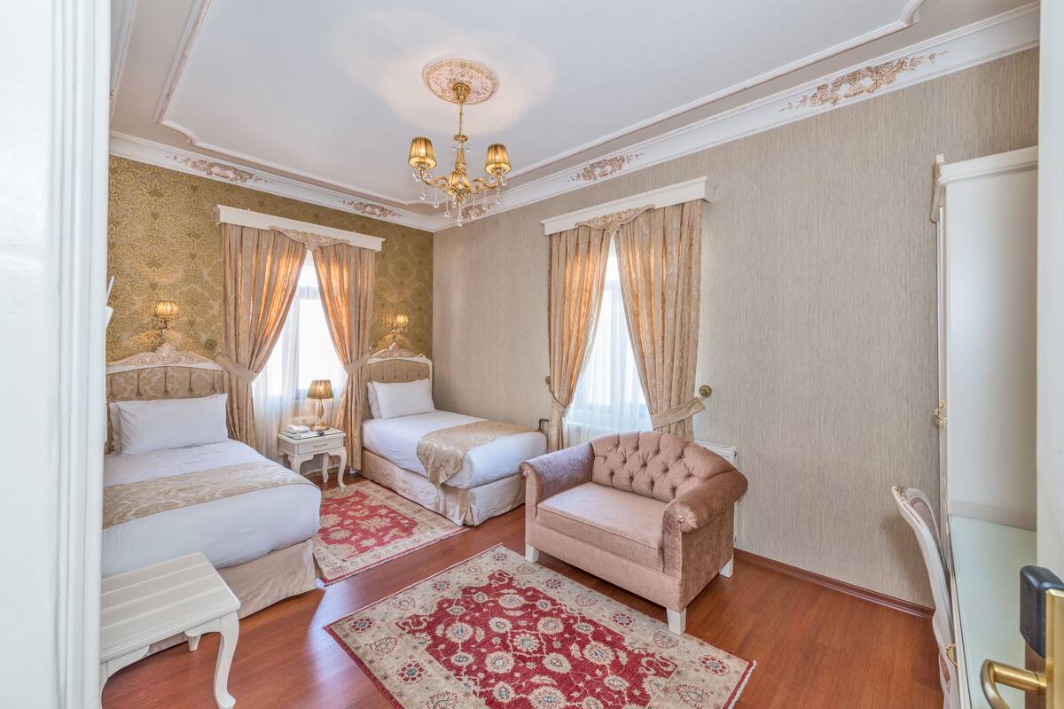 Quarto de hotel com duas camas, área de estar, um candelabro e uma janela grande com cortinas.